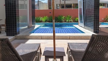 Uma área de piscina com espreguiçadeiras e uma pequena mesa, cercada por paredes revestidas de azulejos e plantas.