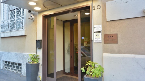Entrada de hotel com porta de vidro, placa indicando 'Hotel Corallo' e duas plantas em vasos na calçada.