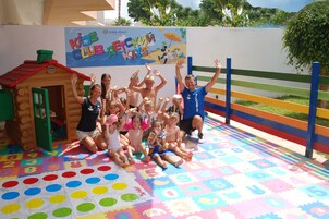 Um grupo de crianças e adultos reunidos em volta de uma casinha de brincar e um tapete colorido com números e letras, em frente a uma parede com um personagem de desenho animado e o texto "Kios Club".