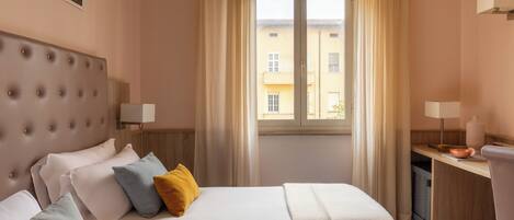 Quarto de hotel com cama, escrivaninha, cadeira, janela com cortinas e vista para prédios.