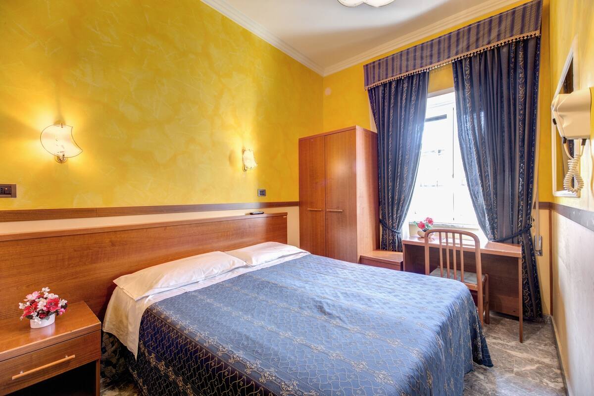 Quarto de hotel com cama, escrivaninha, guarda-roupa e janela com cortinas.