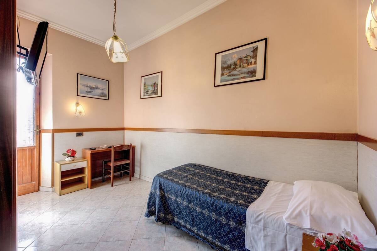 Quarto com cama, escrivaninha e cadeira. Há luminárias fixadas na parede e quadros emoldurados.