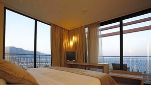 Quarto com cama, televisão, escrivaninha e amplas janelas com vista para o mar.