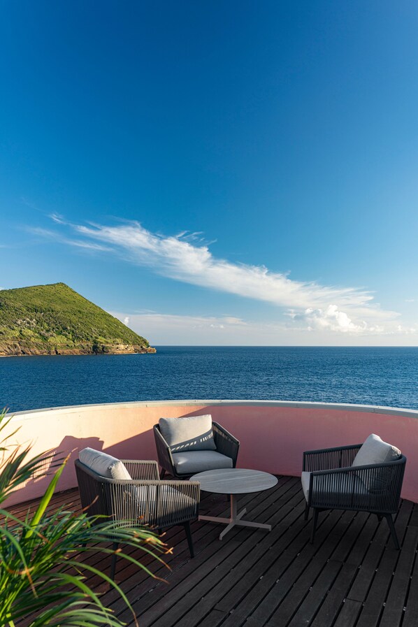 Um terraço com duas cadeiras de vime e uma pequena mesa, com vista para um mar tranquilo e uma colina verde.