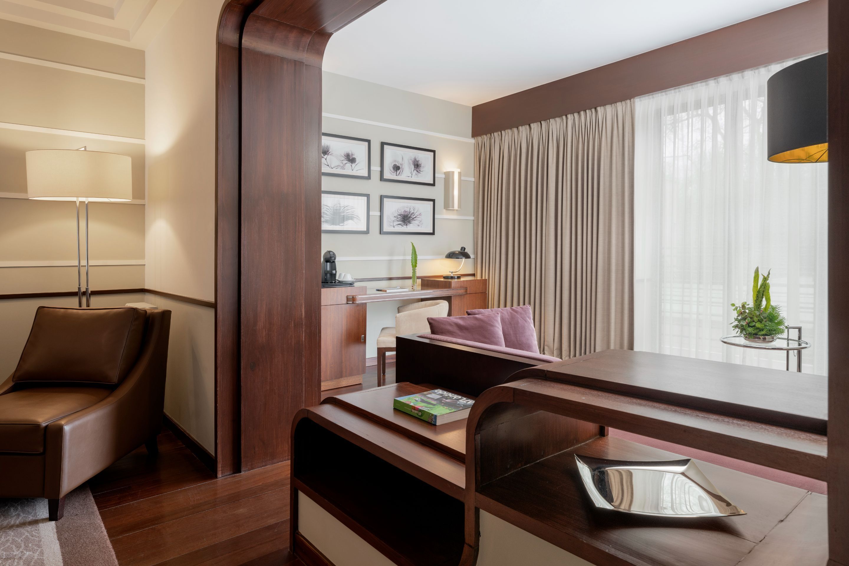 Quarto de hotel moderno com uma escrivaninha de madeira, uma poltrona de couro marrom, uma estante, uma mesa pequena com uma planta e vista para a cidade através de grandes janelas.