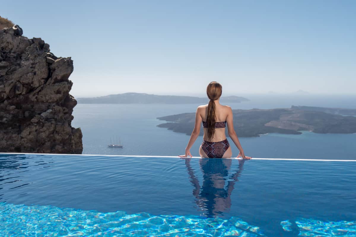 Mulher de biquíni à beira de uma piscina infinita com vista para o mar calmo e a costa rochosa.