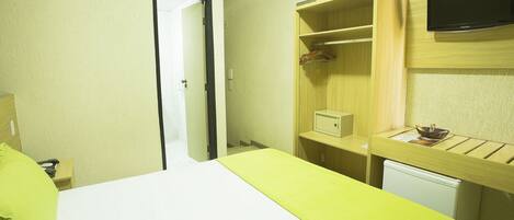 Quarto de hotel com uma cama, uma escrivaninha com cadeira, uma televisão presa na parede e um guarda-roupa.