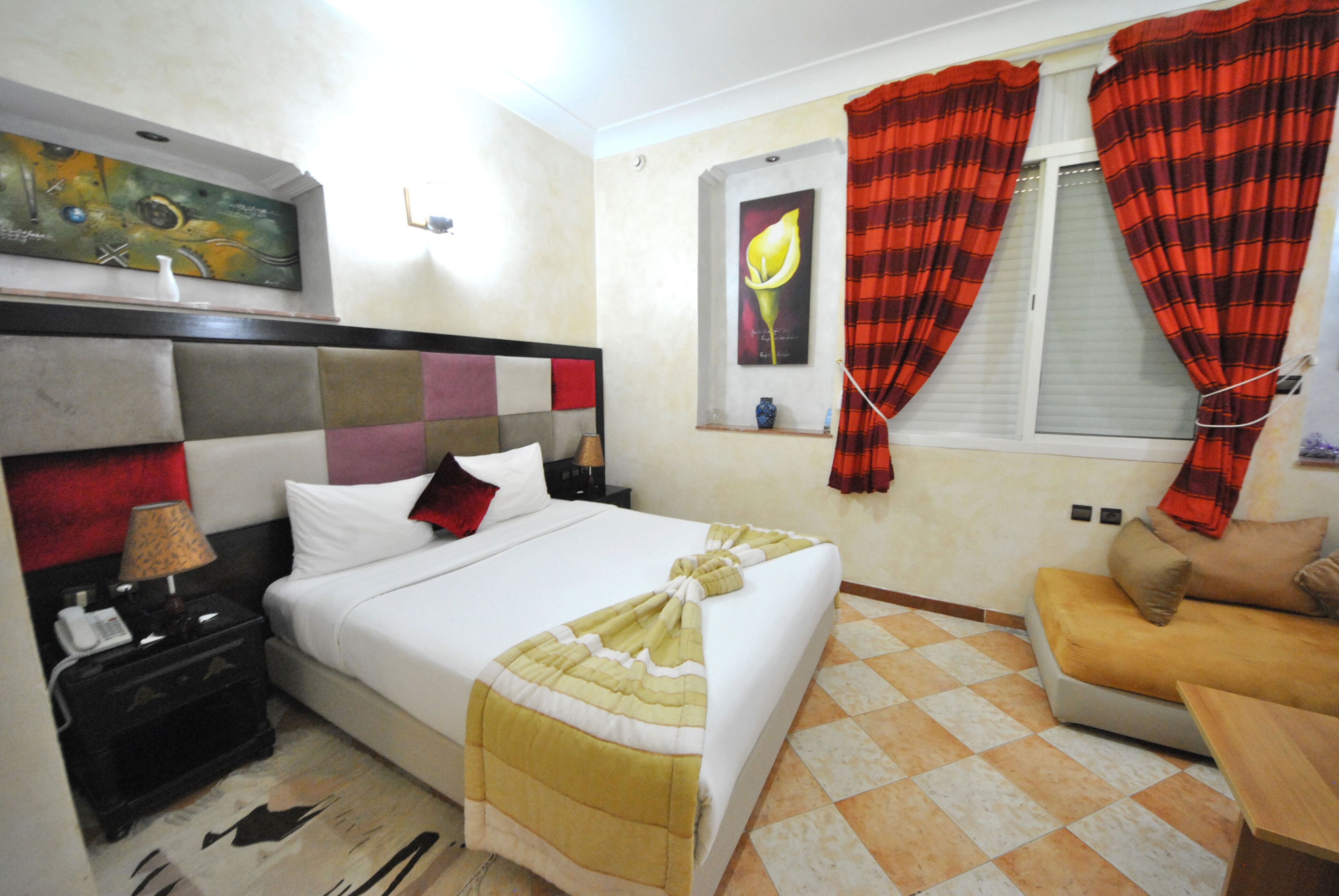 Quarto de hotel com cama, mesa de cabeceira, luminária e janela com cortinas.