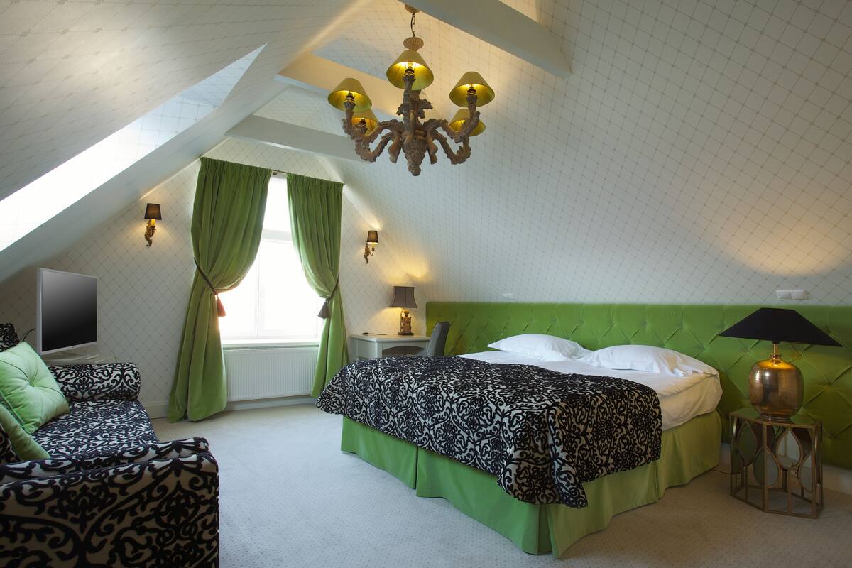 Um quarto com cabeceira estofada em verde, colcha estampada, candelabro e televisão na parede.