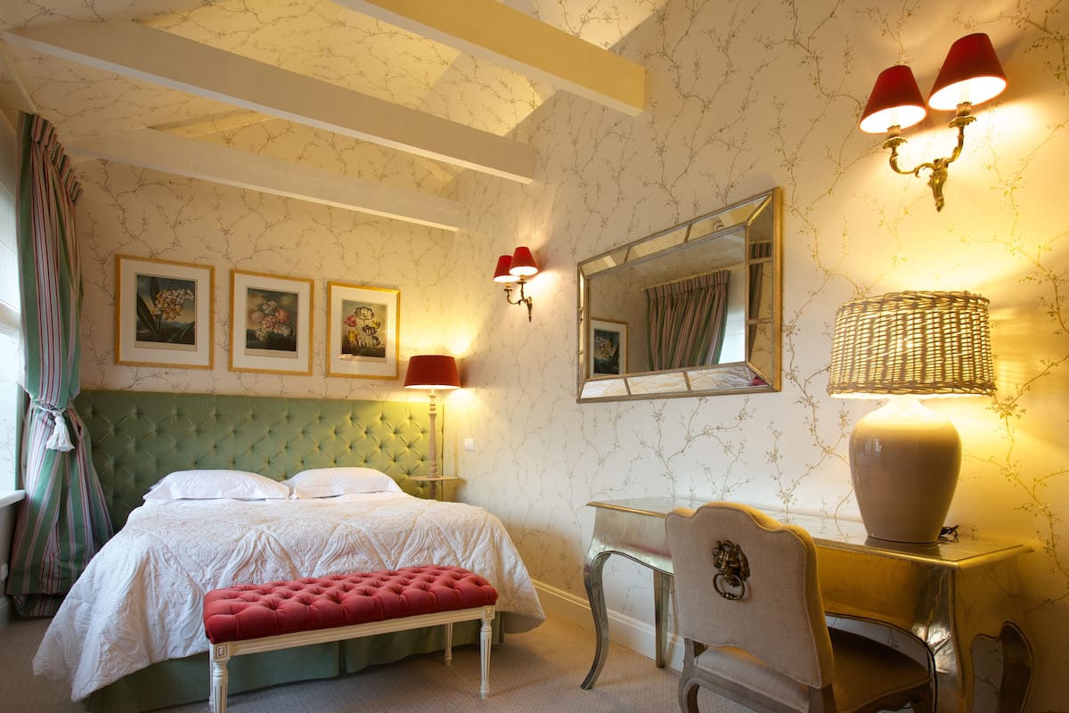Quarto com cabeceira estofada verde, um banquinho vermelho com detalhes em capitonê, uma escrivaninha de madeira e um espelho em cima.