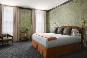 Quarto de hotel com uma cama grande, uma cadeira, uma mesinha e um abajur.