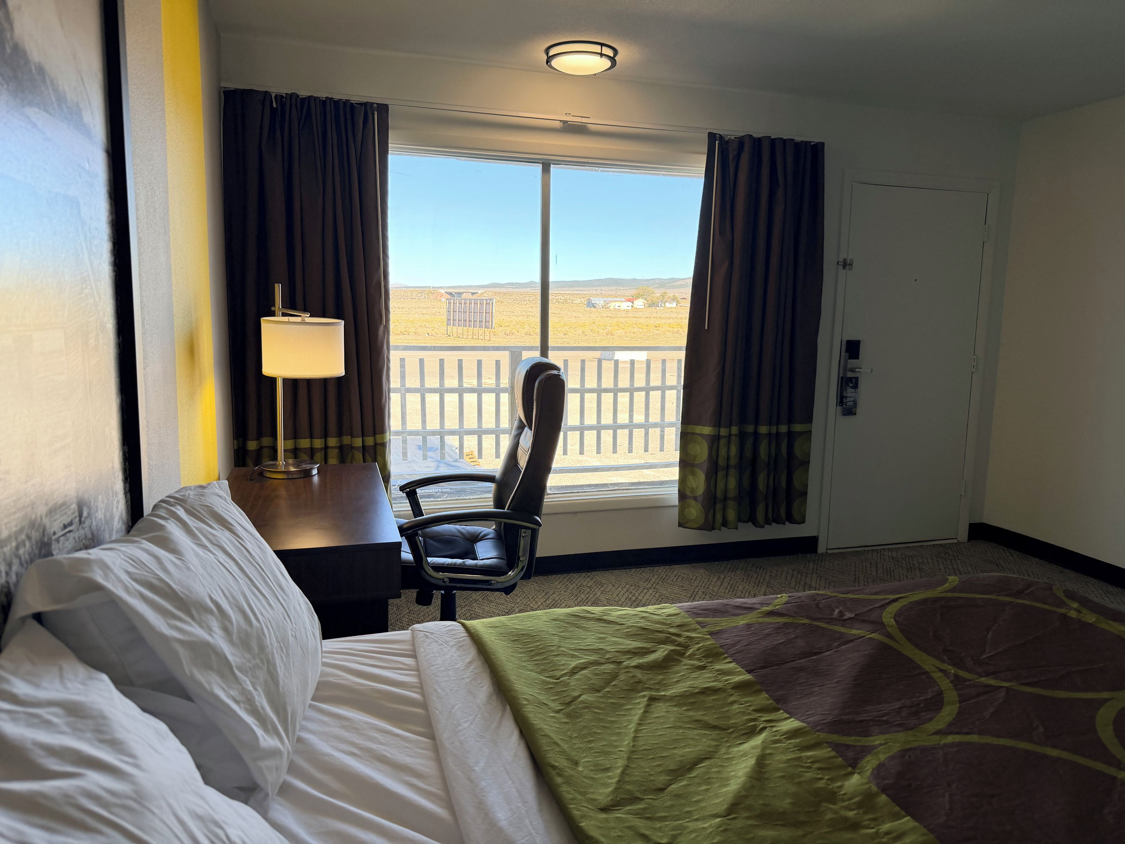 Quarto de hotel com cama, escrivaninha, cadeira, janela com cortinas e porta.