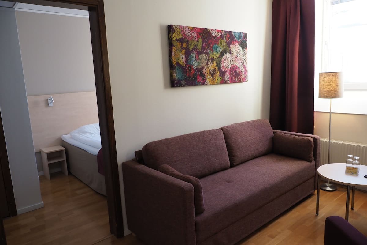 Quarto de hotel moderno com um sofá marrom, uma mesinha de centro com abajur, um quadro abstrato colorido na parede e uma cama com roupa de cama branca.