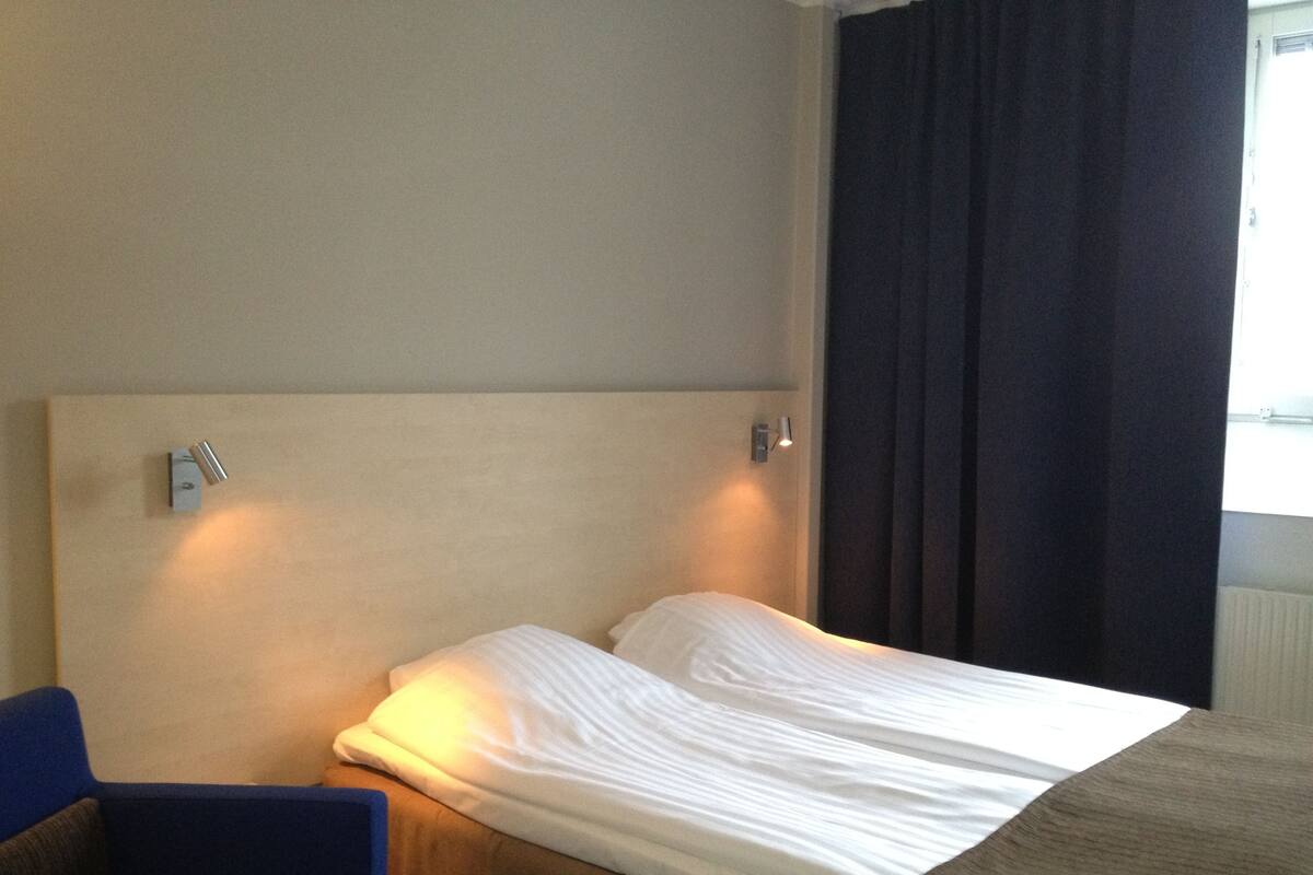 Quarto de hotel com cama, cabeceira, mesa de cabeceira, abajur e janela com cortinas.