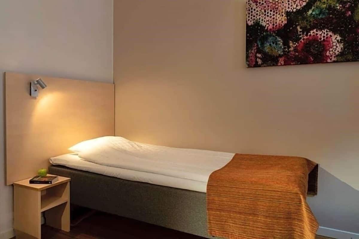 Uma cama de solteiro com cabeceira, uma mesa de cabeceira com um livro e uma maçã, e uma pintura abstrata colorida na parede.