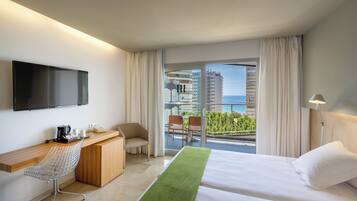 Quarto de hotel com cama, escrivaninha com cadeira, televisão e varanda com vista para o oceano e edifícios.