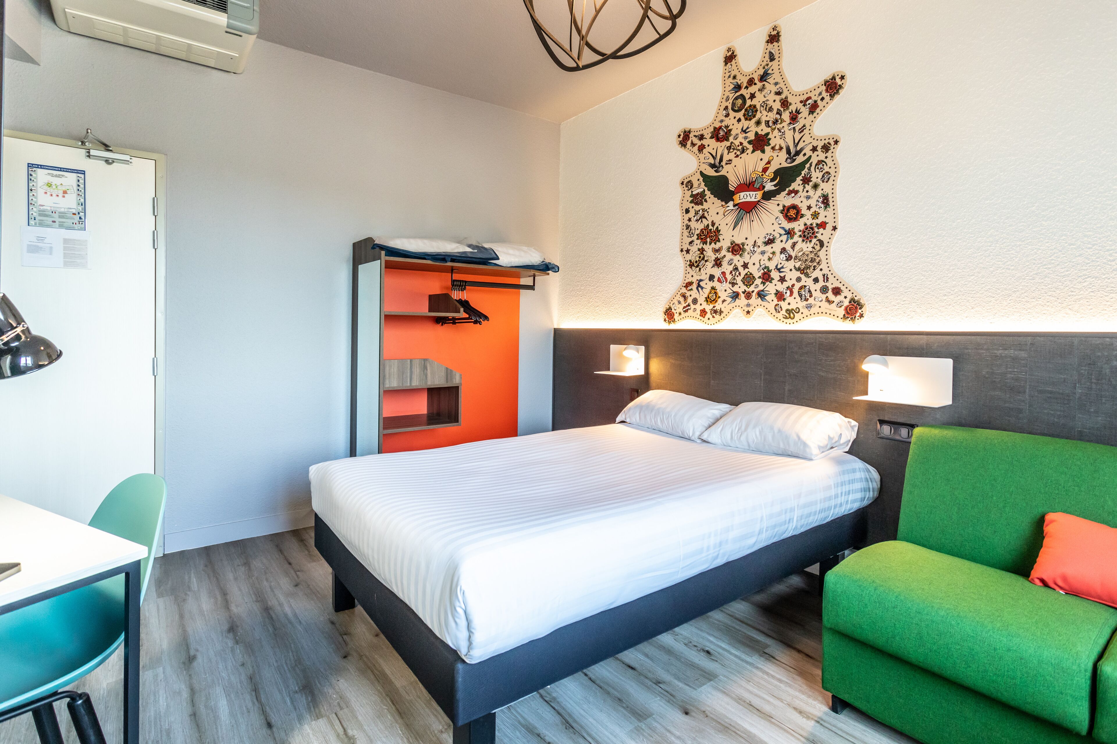 Quarto de hotel com cama, escrivaninha, cadeira, sofá verde e parede com pintura decorativa de um animal.