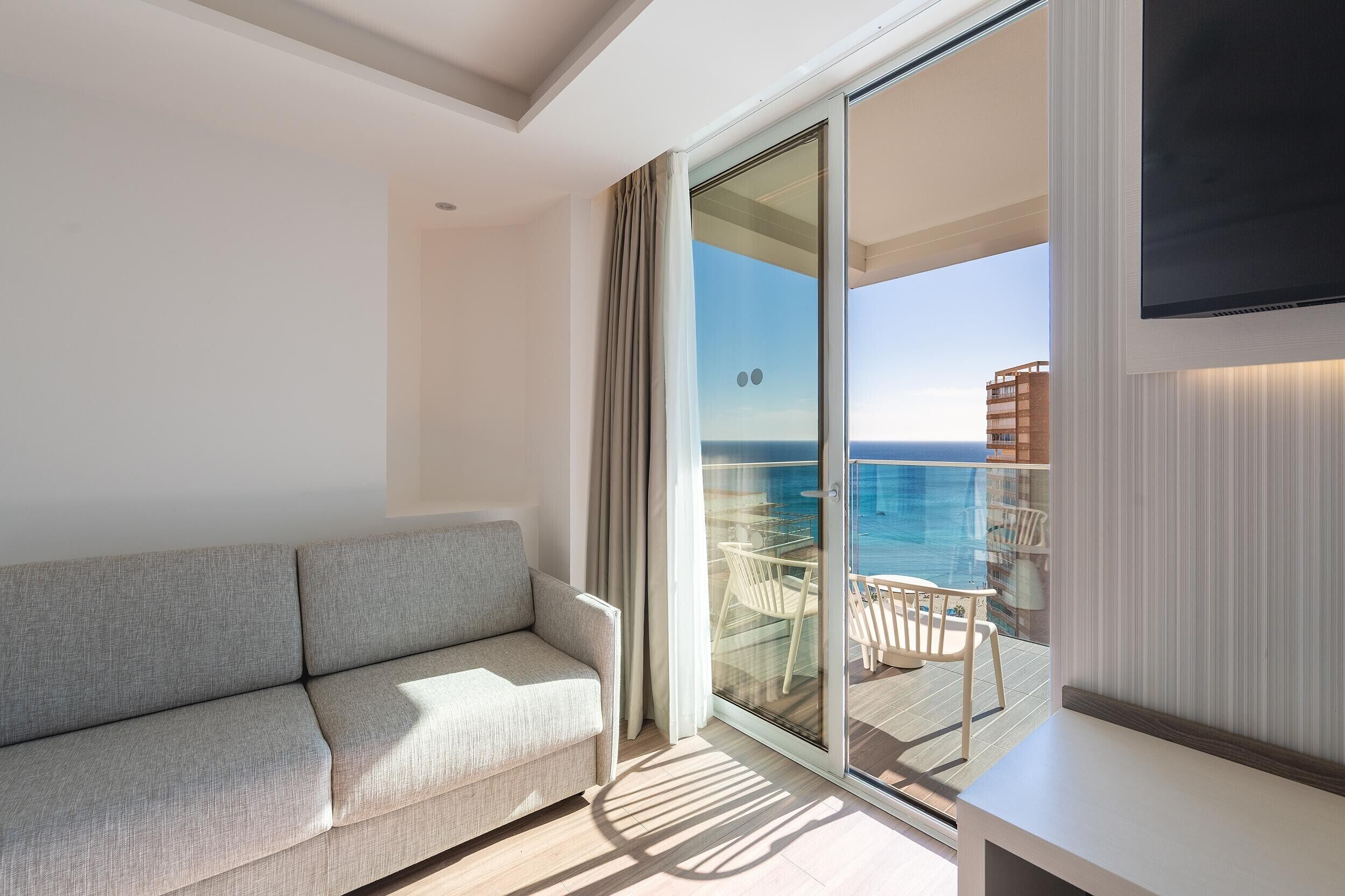 Uma sala de estar moderna com sofá, mesa e porta de vidro que dá para uma varanda com vista para o mar e um edifício.