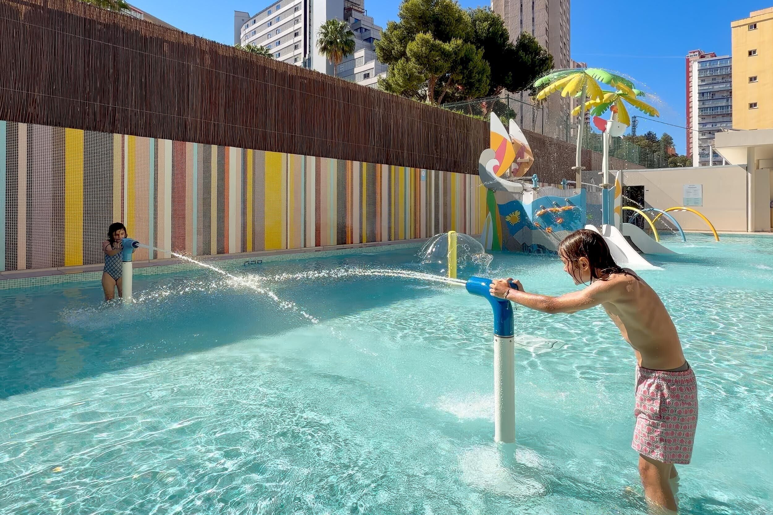 Uma área de piscina com estrutura de recreação aquática, cerca com listras verticais coloridas e edifícios ao fundo.