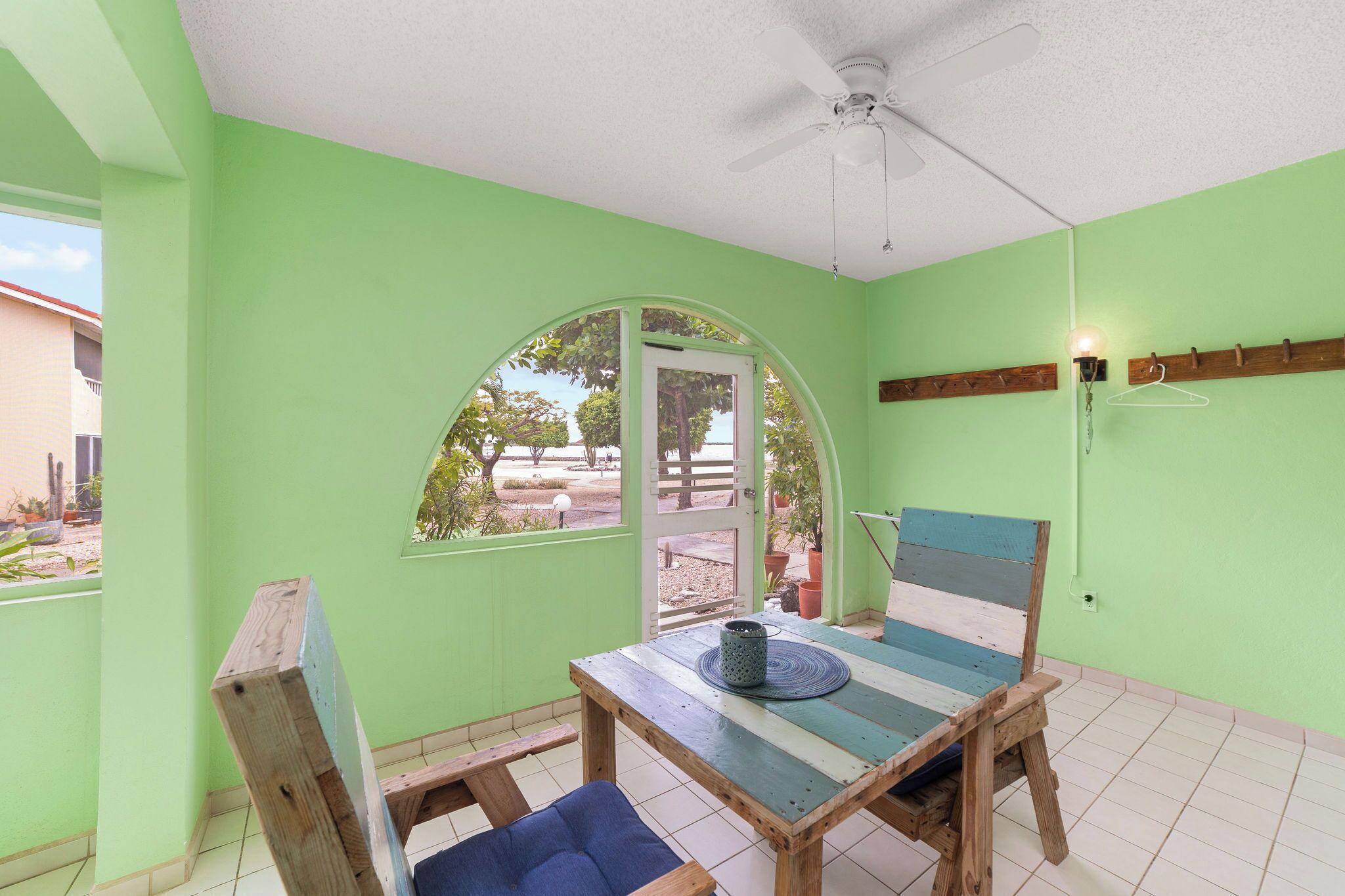 Quarto com parede verde, mesa de madeira e cadeira listrada, ventilador de teto e janela com vista para o exterior.