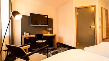 Quarto de hotel com cama, escrivaninha com cadeira, televisão e porta.