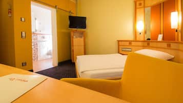 Quarto de hotel com cama, televisão fixada na parede, armário de madeira e banheiro visível através de uma porta aberta.