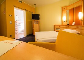 Quarto de hotel com cama, televisão fixada na parede, armário de madeira e banheiro visível através de uma porta aberta.