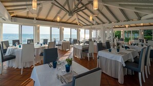 Restaurante com mesas preparadas para refeições, grandes janelas com vista para o oceano e teto alto com vigas aparentes.