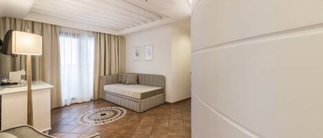 Quarto de hotel com sofá, mesa pequena, luminária e janela com cortinas.