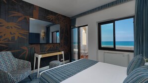Quarto de hotel moderno com uma cama grande, uma mesa de trabalho e uma cadeira. Há uma janela ampla com vista para a praia e o mar.