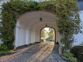 Uma passagem arqueada com piso de pedras, rodeada por vegetação exuberante e paredes brancas.