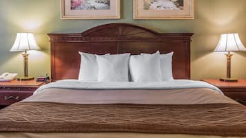 Cama bem arrumada com almofadas brancas e manta marrom, acompanhada por duas mesas de cabeceira com abajures, em frente a uma parede verde com duas imagens emolduradas.