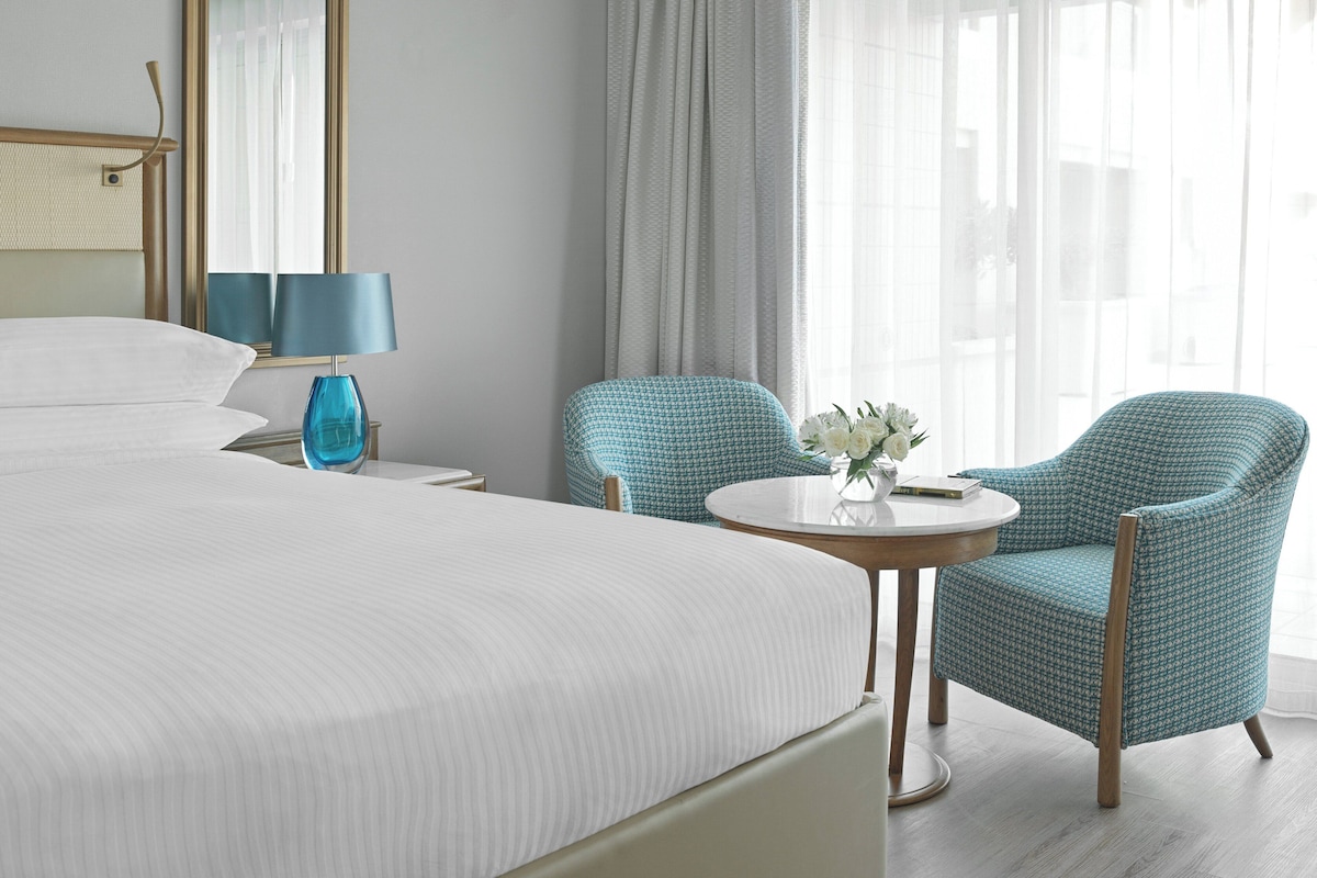 Quarto de hotel com uma cama grande, duas cadeiras, uma mesa pequena e um abajur.