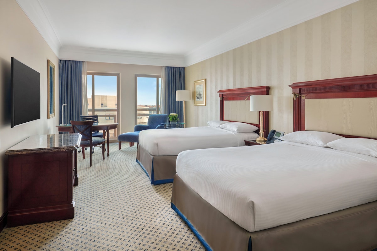 Quarto de hotel com duas camas, uma mesa, uma cadeira, uma televisão e uma janela com cortinas.