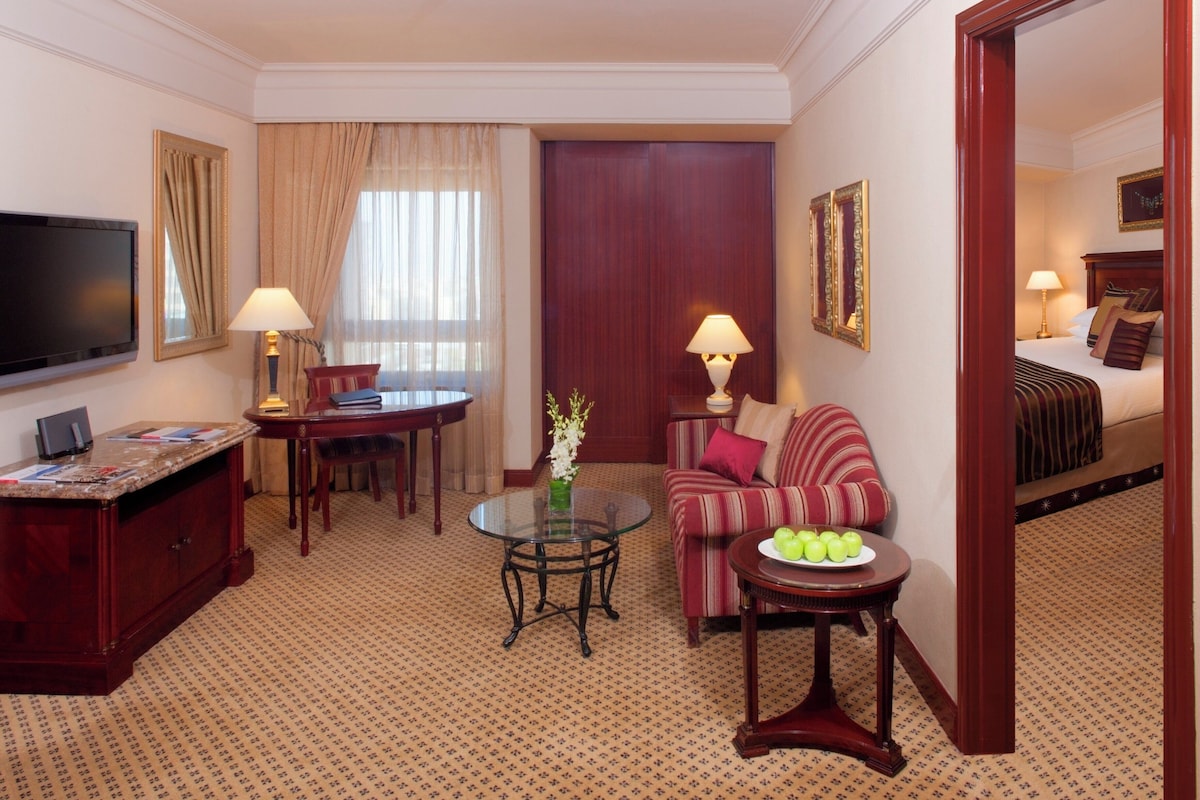Quarto de hotel com televisão, escrivaninha, mesa de jantar, sofá, mesinha de centro e cama.