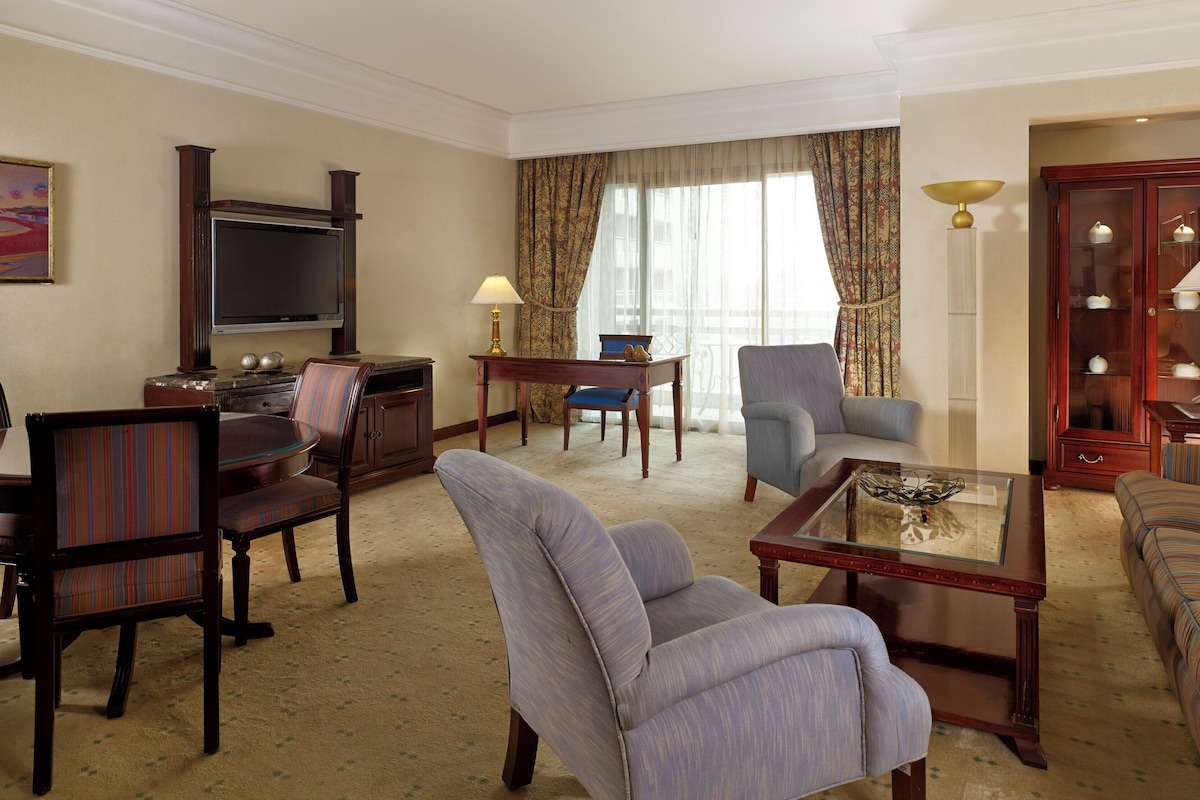 Quarto de hotel com televisão, área de estar e uma janela grande com cortinas.