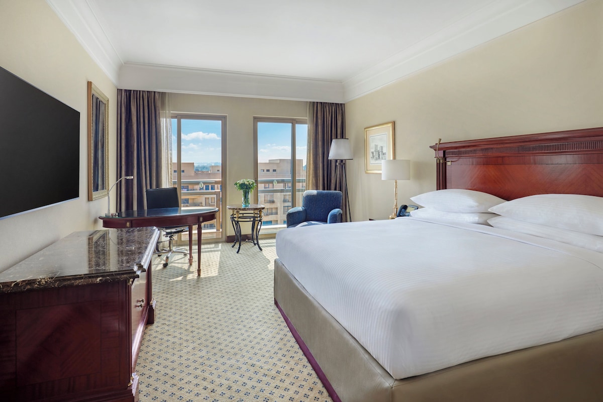Quarto de hotel com uma cama grande, uma escrivaninha, uma cadeira, uma mesa pequena e uma varanda com vista para a cidade.