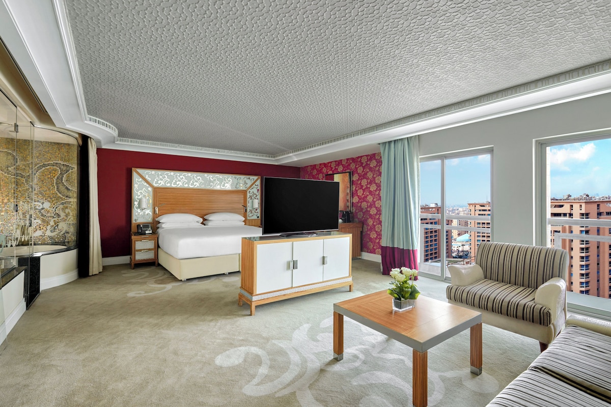 Quarto de hotel com cama, televisão, sofá, mesa de centro e vista para a cidade.
