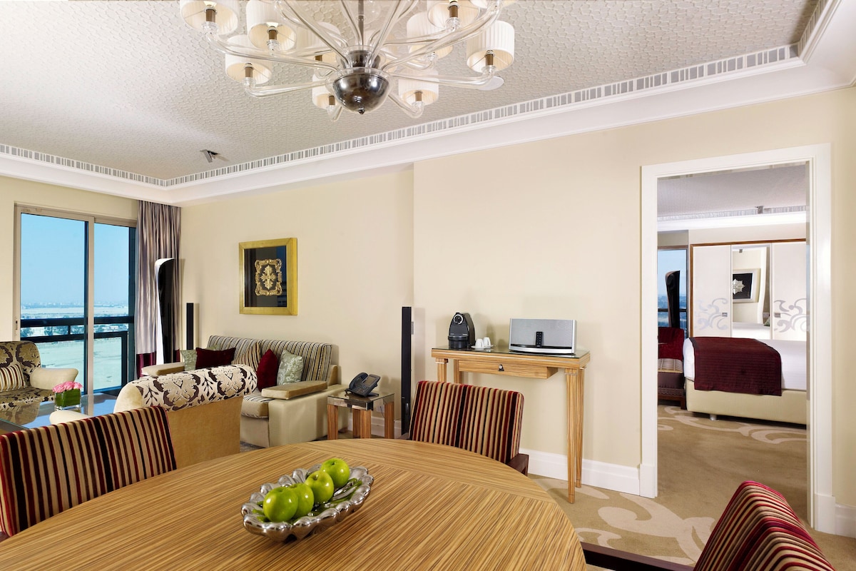 Quarto de hotel com área de refeições, sofás, televisão e vista para a cidade.
