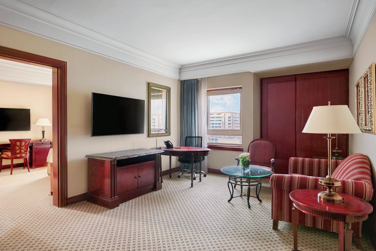 Quarto de hotel com escrivaninha, televisão, sofá, poltrona, mesa e janela com vista para edifícios.