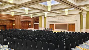 Sala de conferências com fileiras de cadeiras pretas, carpete estampado e teto alto com lustres.