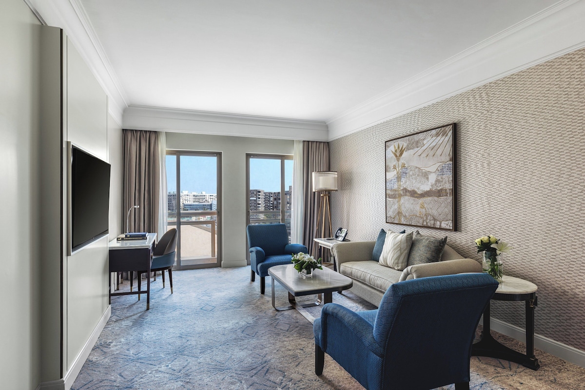 Quarto de hotel com área de estar, televisão de tela plana, escrivaninha com luminária e uma grande janela com vista para a cidade.