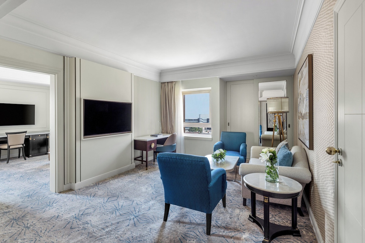 Quarto de hotel com televisão de tela plana, escrivaninha, poltrona azul, sofá e mesa de centro.