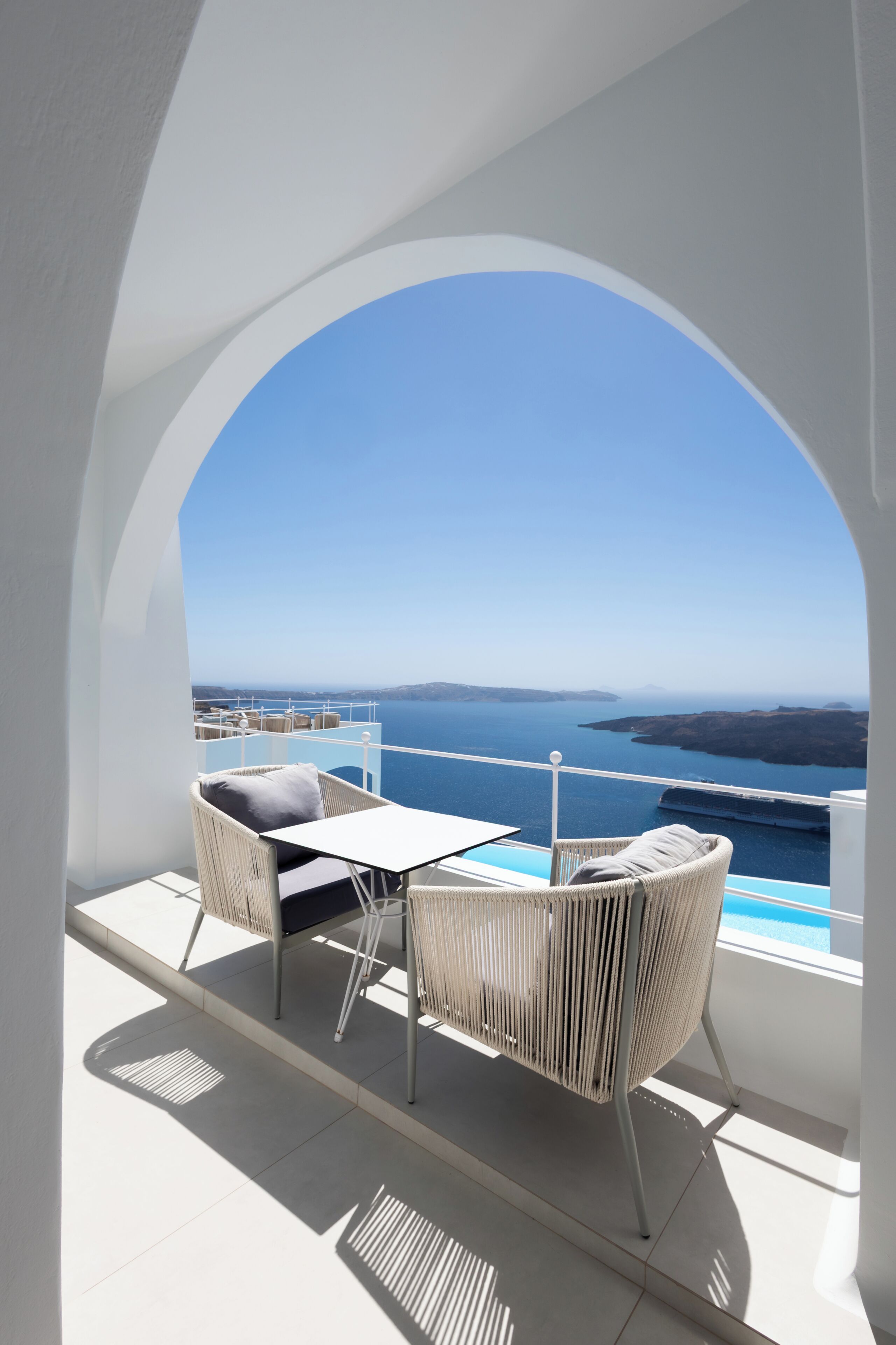 Uma varanda com arco branco, duas cadeiras de vime, uma pequena mesa e vista para o mar e terra ao longe.