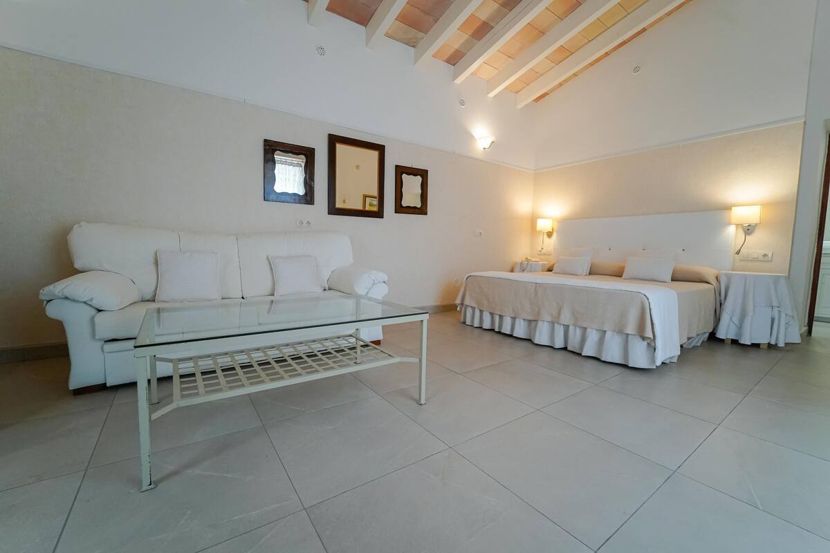 Quarto de hotel moderno com um sofá branco, uma mesa de centro de vidro, uma cama com edredão branco e duas luminárias fixadas na parede.