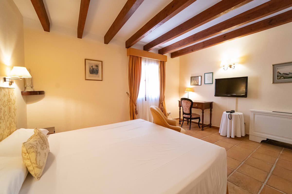 Quarto de hotel com uma cama grande, uma cadeira, uma mesinha, uma televisão e um radiador.