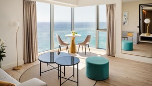 Quarto de hotel moderno com uma grande janela que oferece vista para o oceano. Há uma mesa redonda com cadeiras, um sofá e uma pequena mesa redonda com uma luminária.