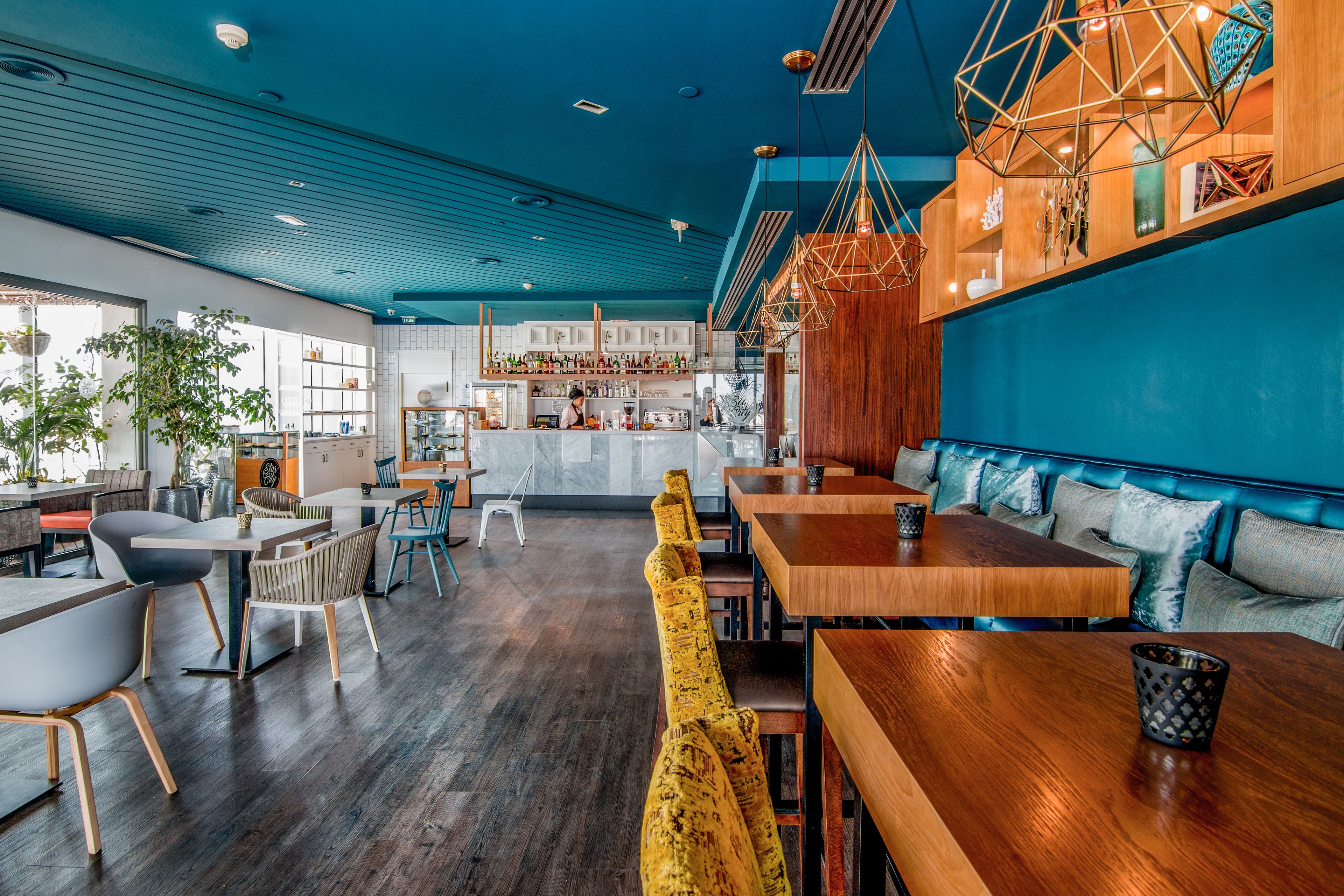 Um restaurante moderno com mesas e bancos de madeira, uma área de bar e uma cozinha ao fundo.