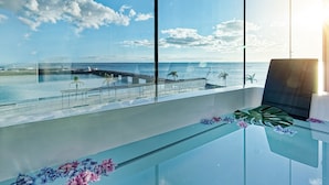 Quarto de hotel moderno com uma grande janela de vidro com vista para uma praia e oceano.