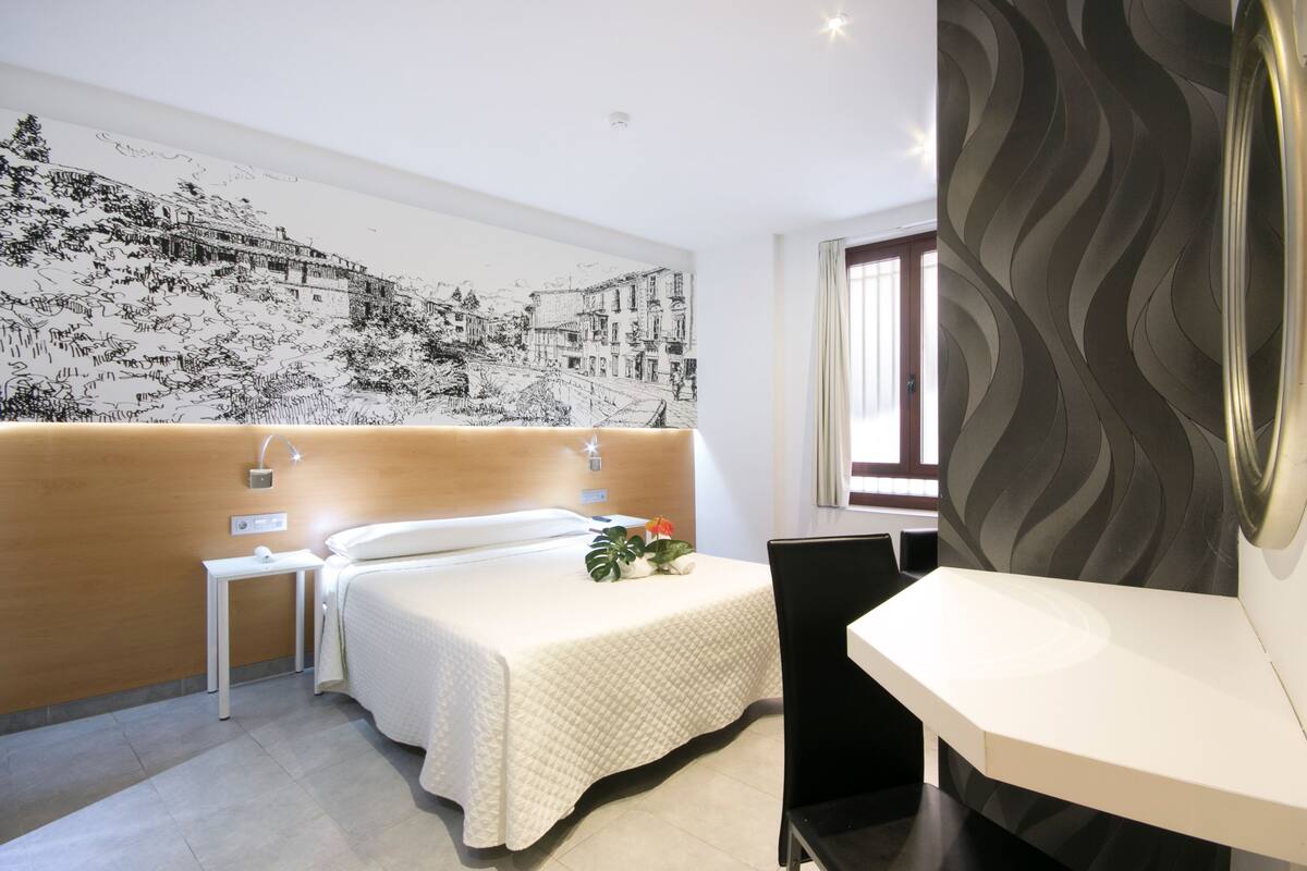 Quarto de hotel com cama, mesas de cabeceira, luminária na parede, uma pequena planta e uma janela grande.